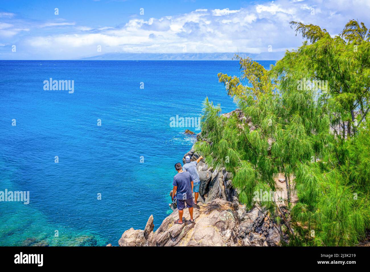 Two male tourists stand on the top of a rugged cliff looking out to the ...