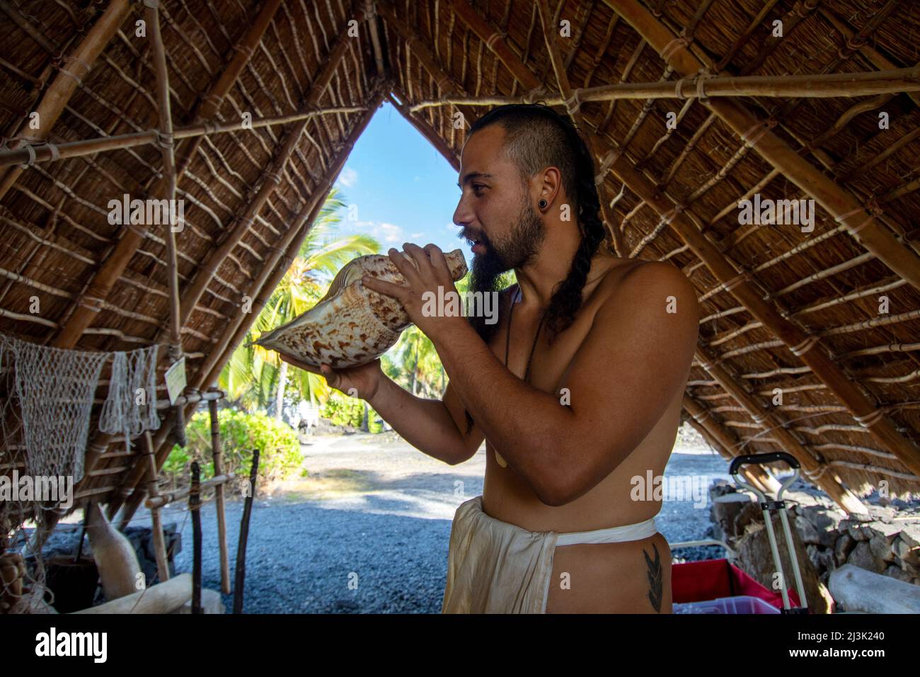 Man in a malo (loincloth) blows into a conch shell. He is the park ...