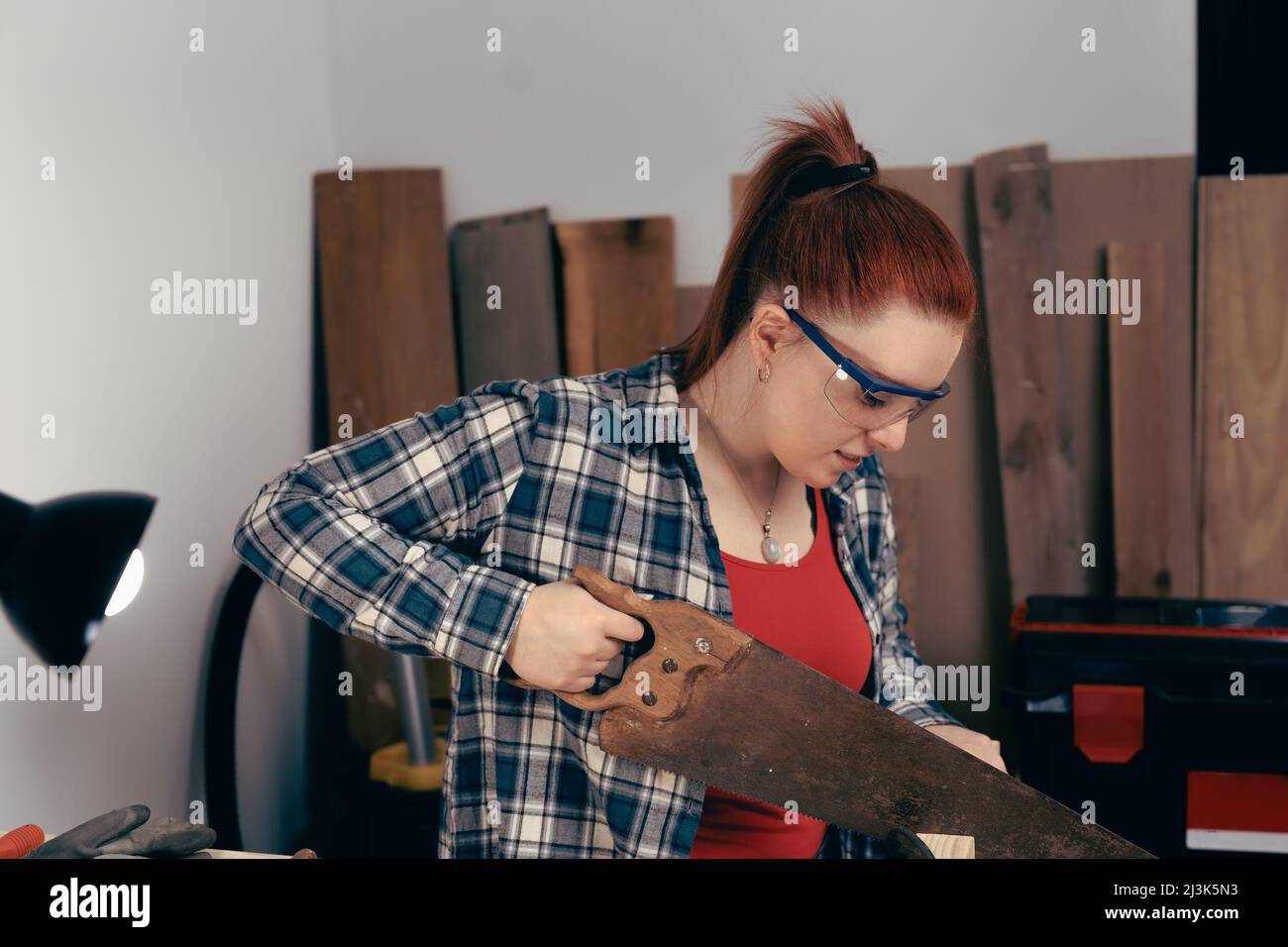 Young red-haired woman, carpenter, sawing a wooden board with a saw, in ...