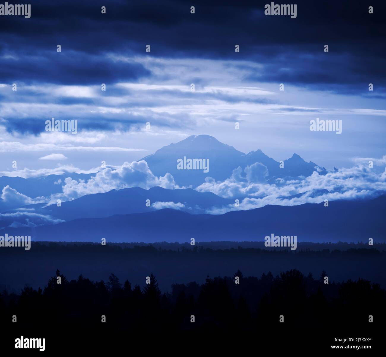 Peaks of Mount Baker with clouds and silhouetted forests in the valley ...