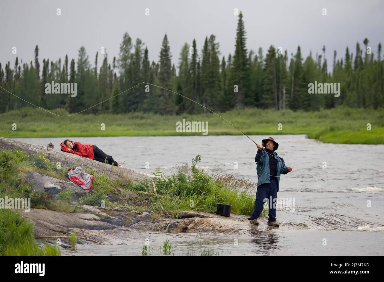 A woman lounges on a rock while her friend casts his fly rod.; Winisk ...