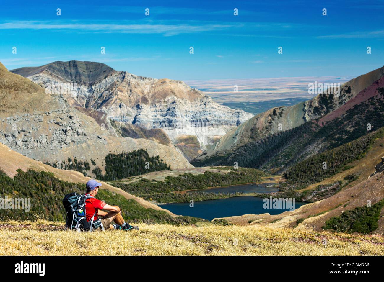 Male hiker relaxing on grassy mountain ridge overlooking an alpine lake ...