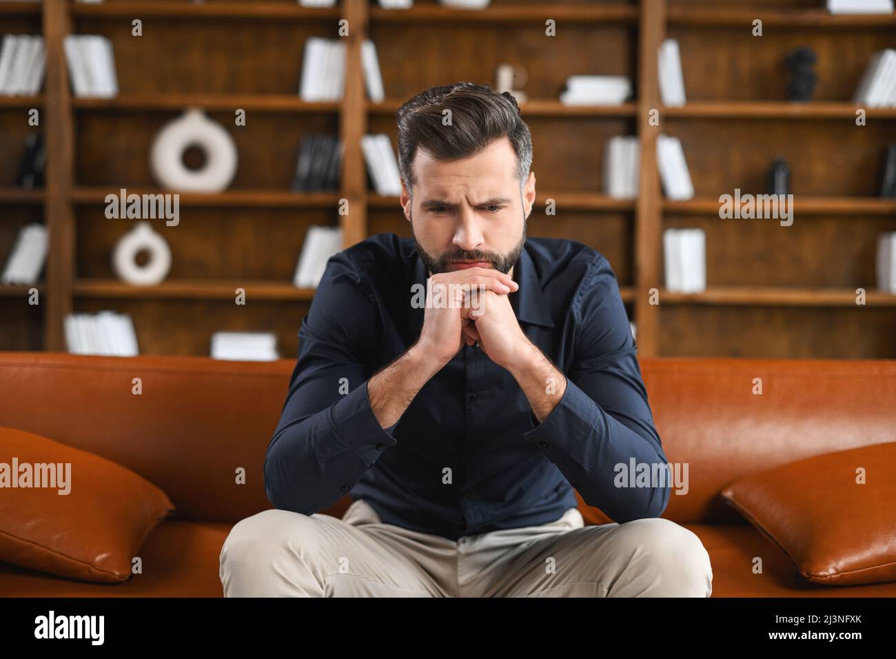 Pensive caucasian guy sitting on the sofa at home or at the office ...