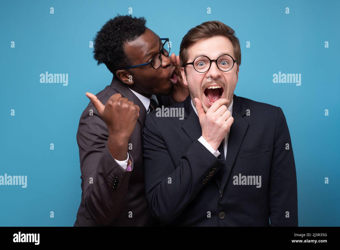 Two shocked young men coworkers whisper gossip and tell secret behind ...