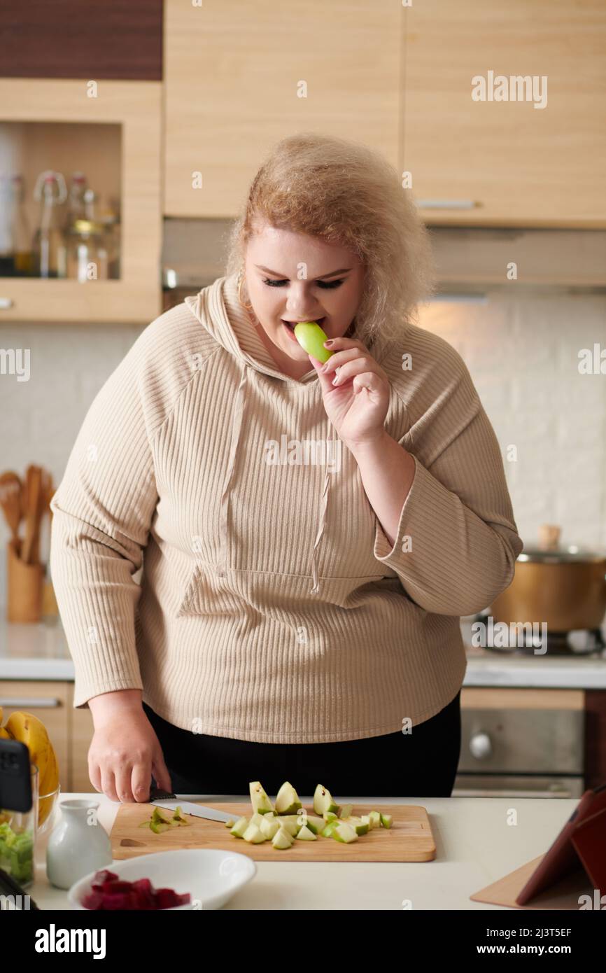Curvy young woman tasting green apple she is cutting for fruit salad at ...