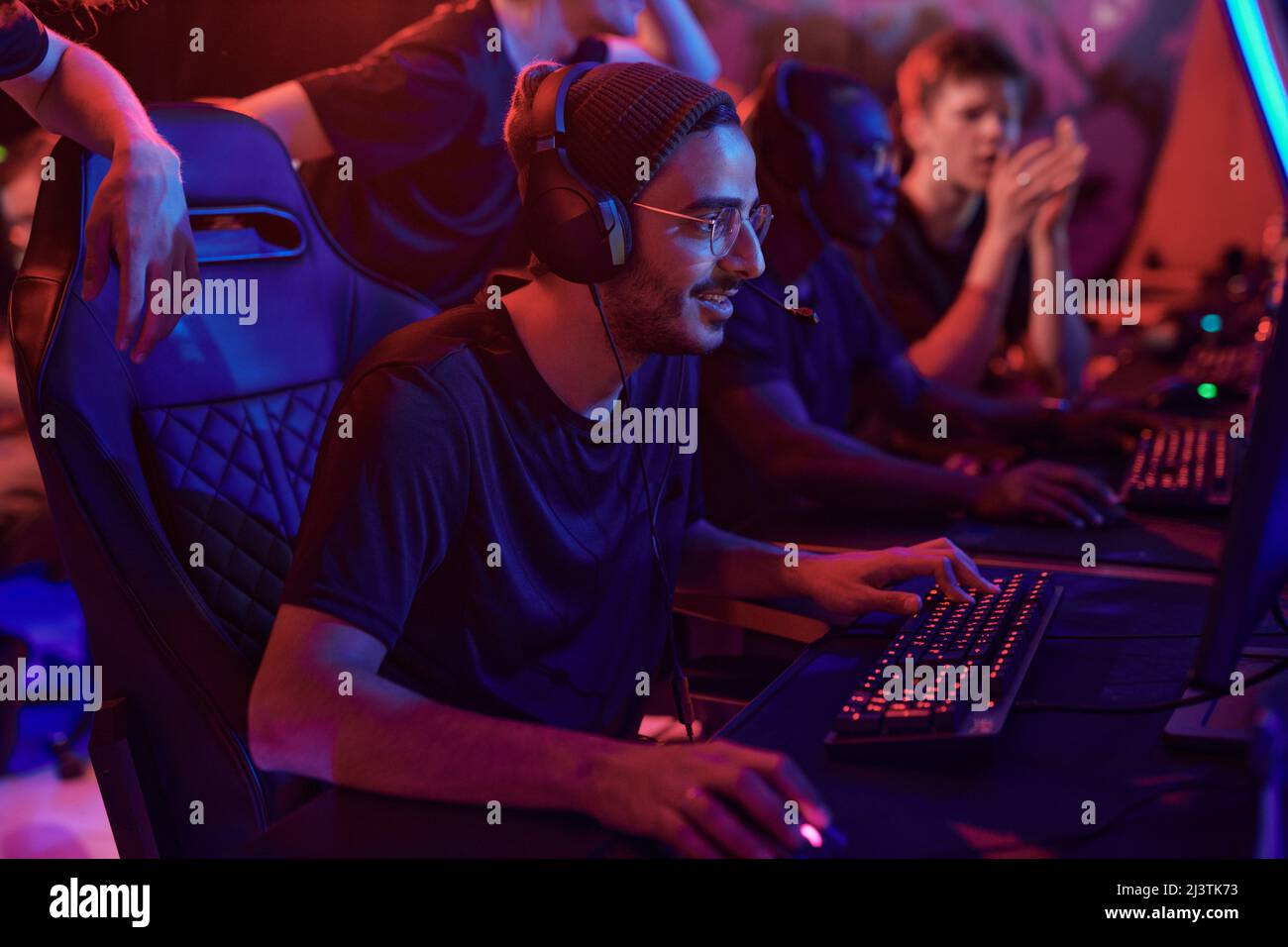 Smiling young professional gamer in hat and eyeglasses using computer ...