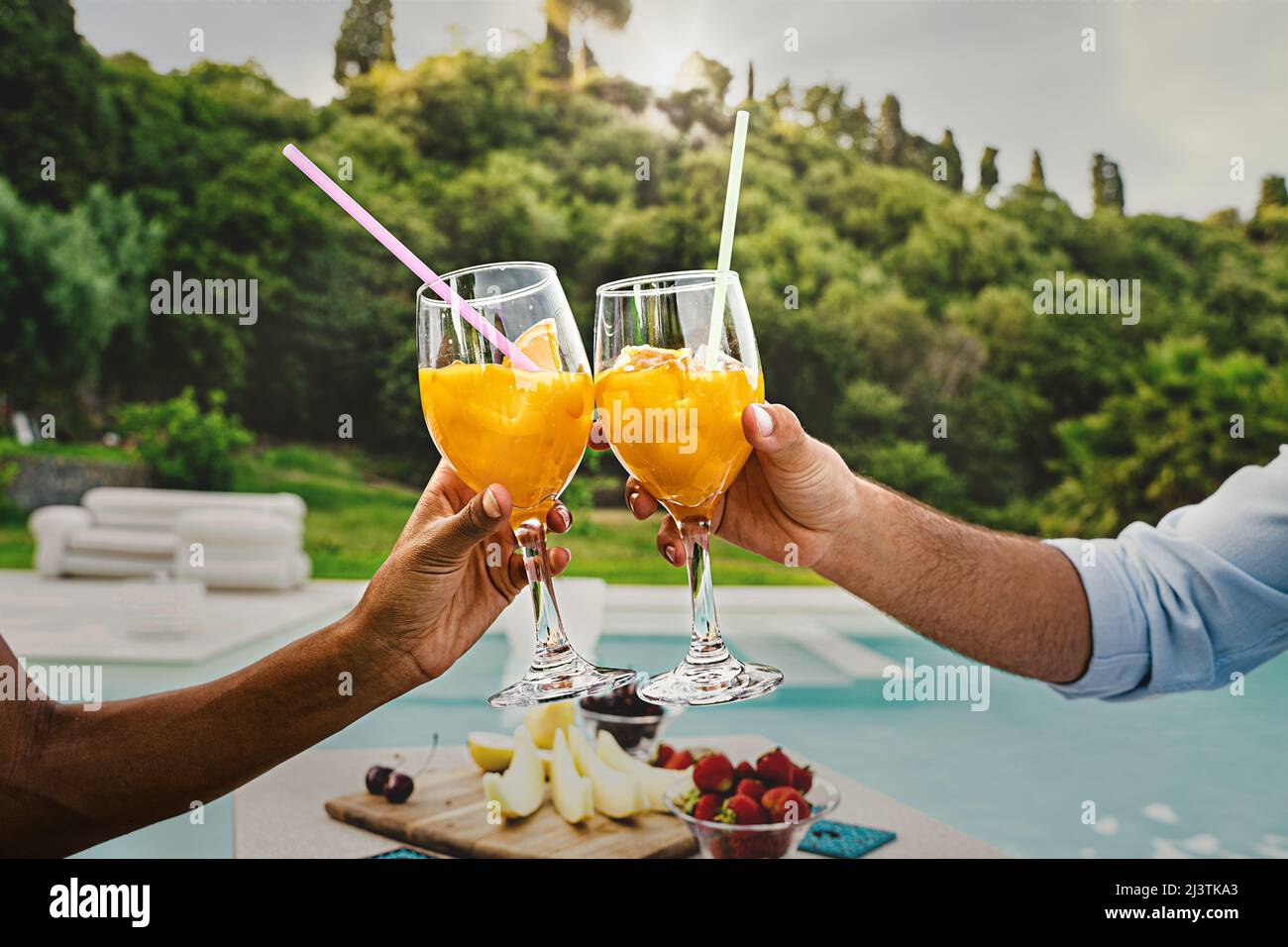 Hands clinking orange cocktail glasses against a swimming pool ...