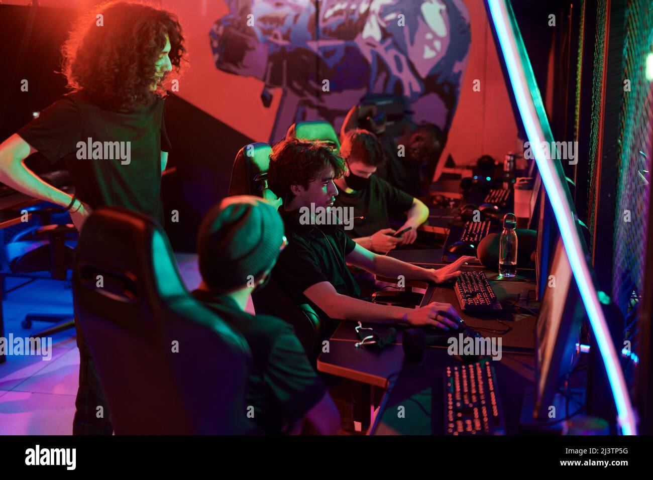 Young curly-haired esports coach standing behind back of gamer and ...