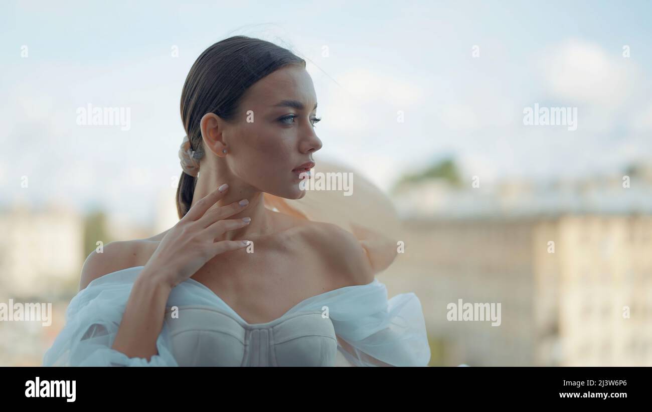 Young beautiful bride in a white wedding dress standing on a balcony on blurred city background ...