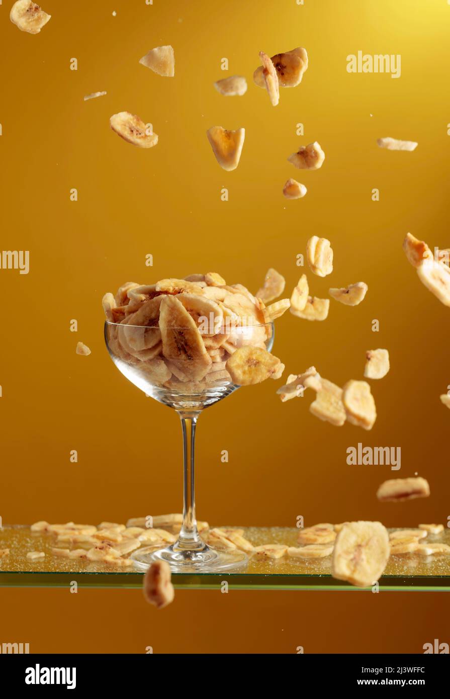 Banana chips fall in a glass bowl. Yellow background with copy space ...