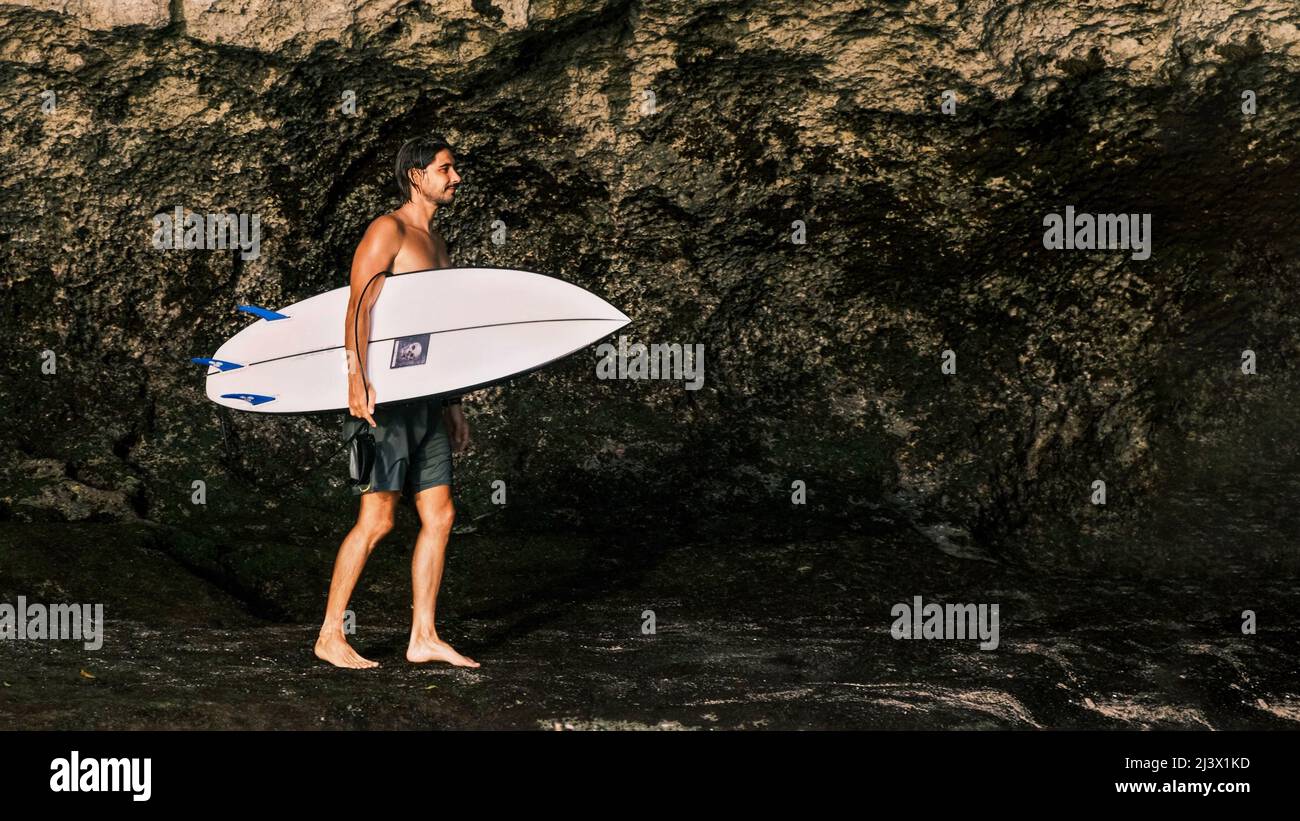 Surfer. Bearded male millennial with bare torso walks along rocks with ...