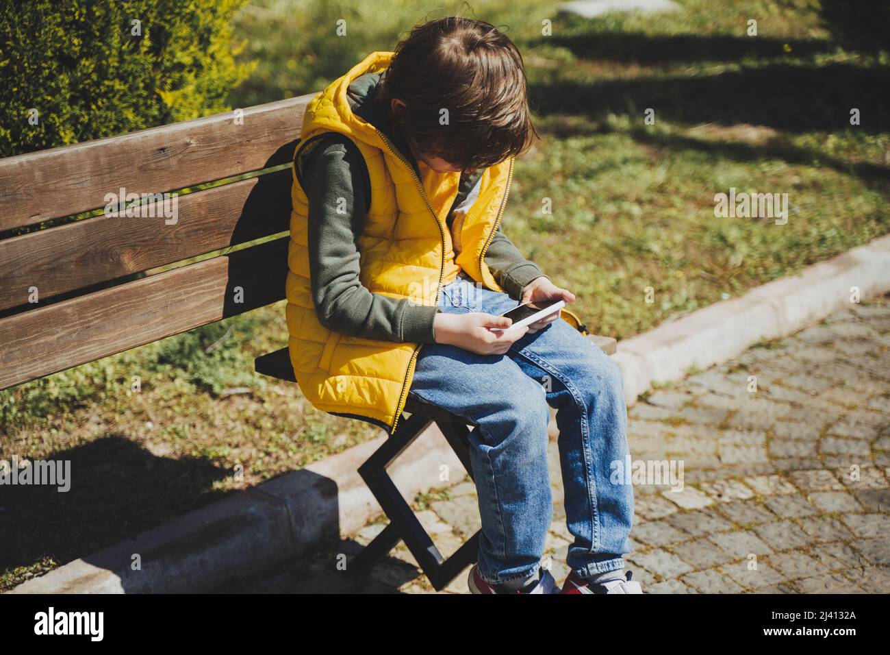 Schoolboy plays game his cell phone sitting on bench in the city park ...
