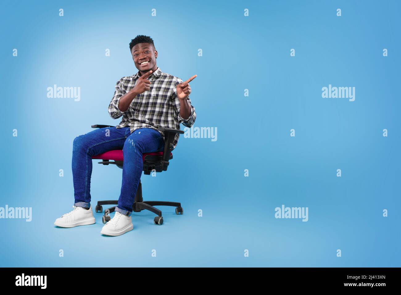 Excited young black man sitting on chair, pointing two fingers aside ...