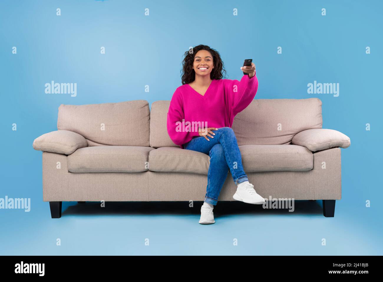 Young African American lady with remote control sitting on couch and ...