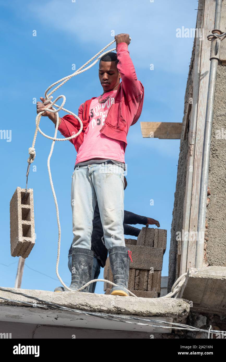 A young Cuban man works as an assistant for a bricklayer. He is lifting ...