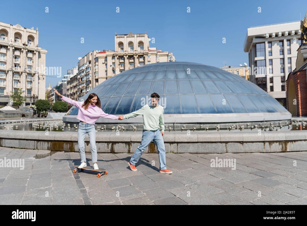 Positive woman riding penny board and holding hands with man on urban ...