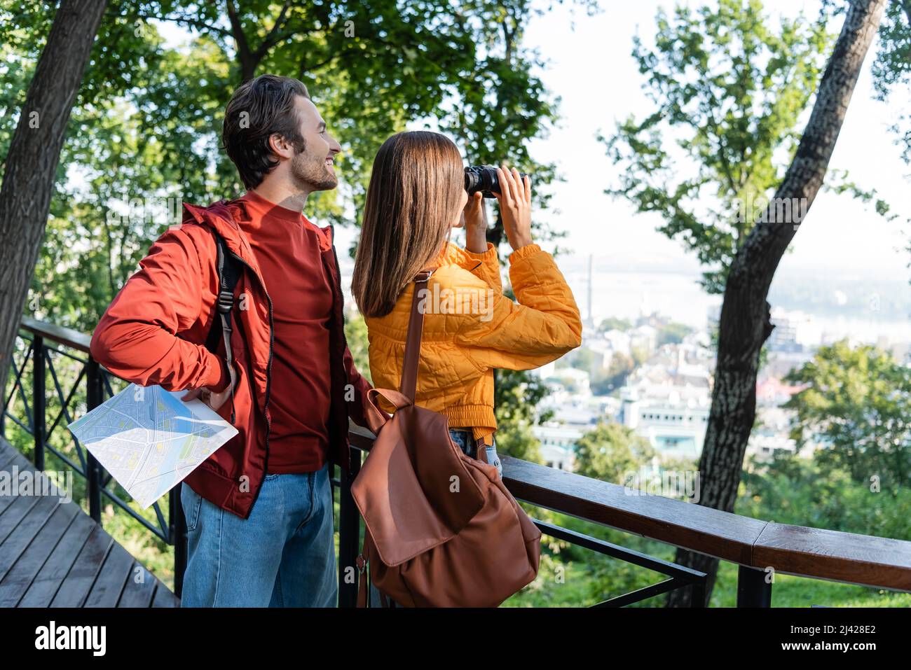 Smiling tourist holding map near girlfriend with binoculars in city ...