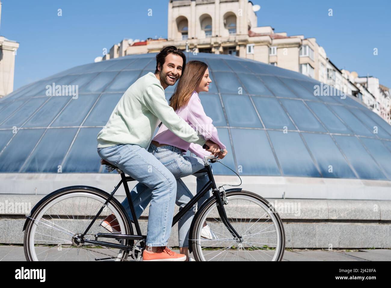 Smiling man looking at camera while cycling with girlfriend on urban ...