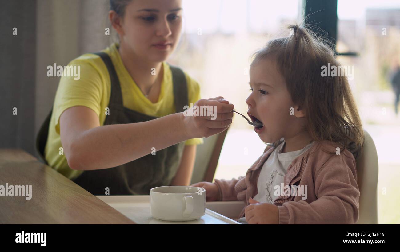 Closeup mom feed young baby in white feeding up high chair, kid is ...