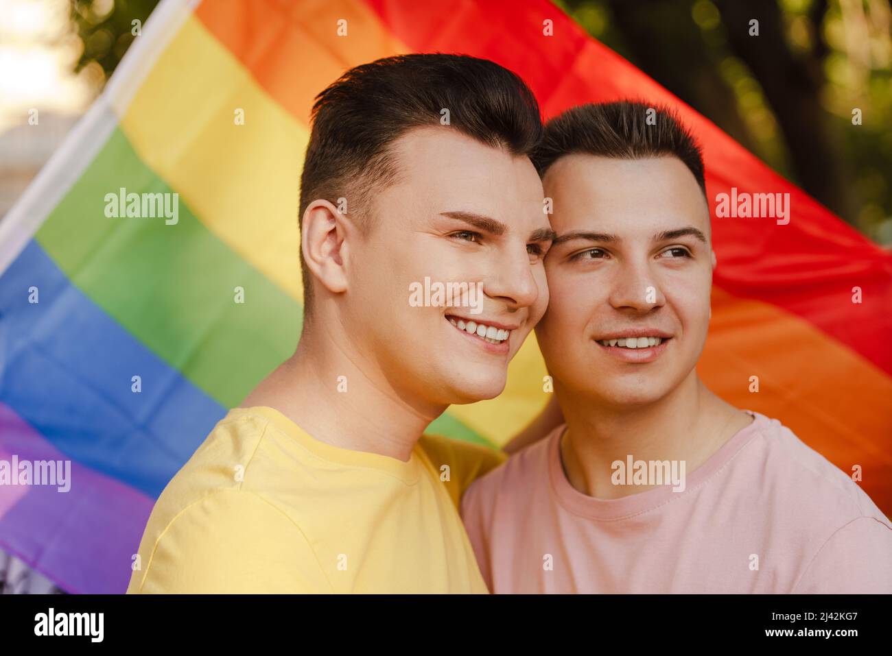 Young gay couple with rainbow flag smiling during pride parade on city ...