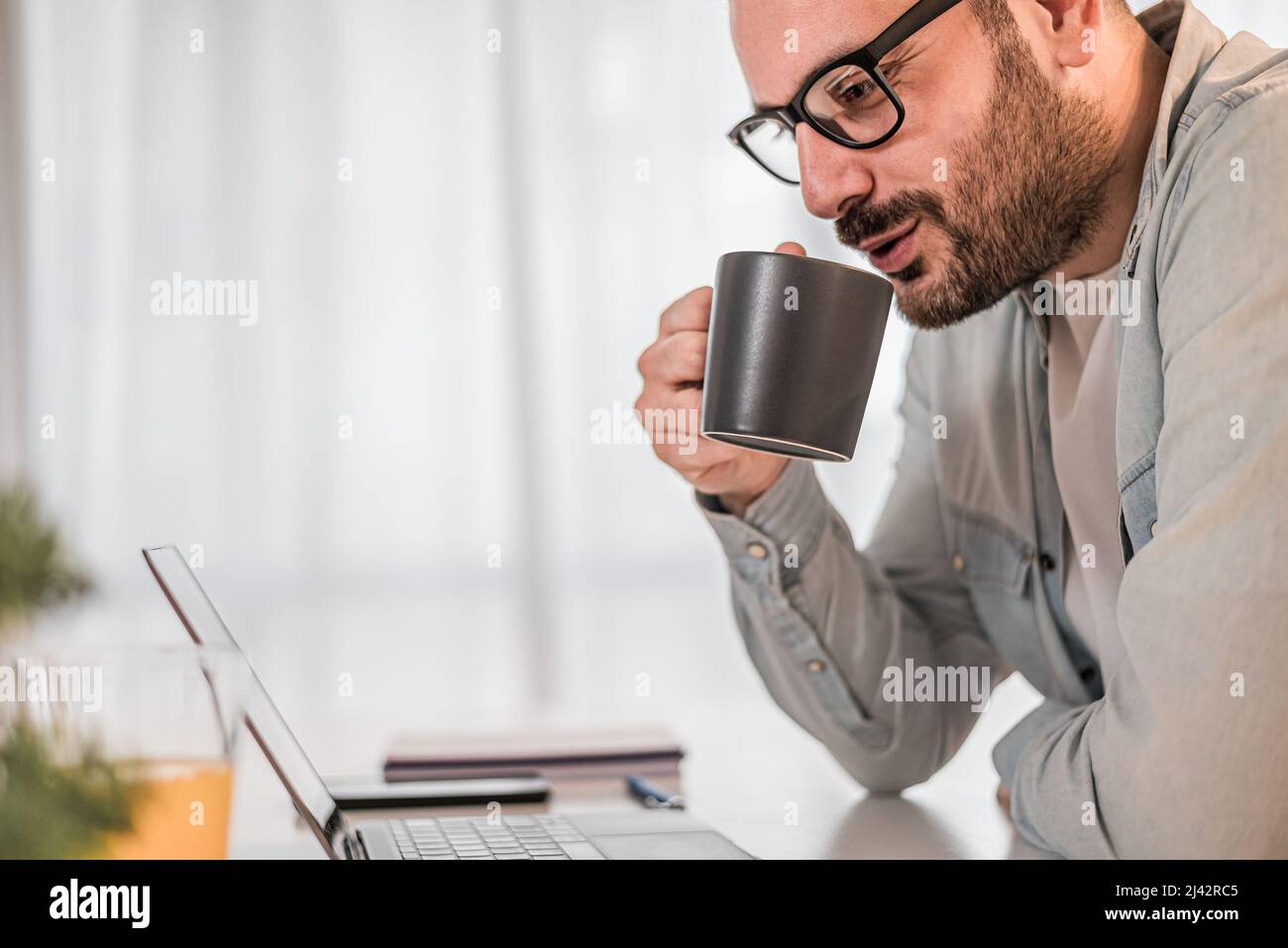 Calm adult man, bringing the cup close to his mouth, glancing at laptop ...