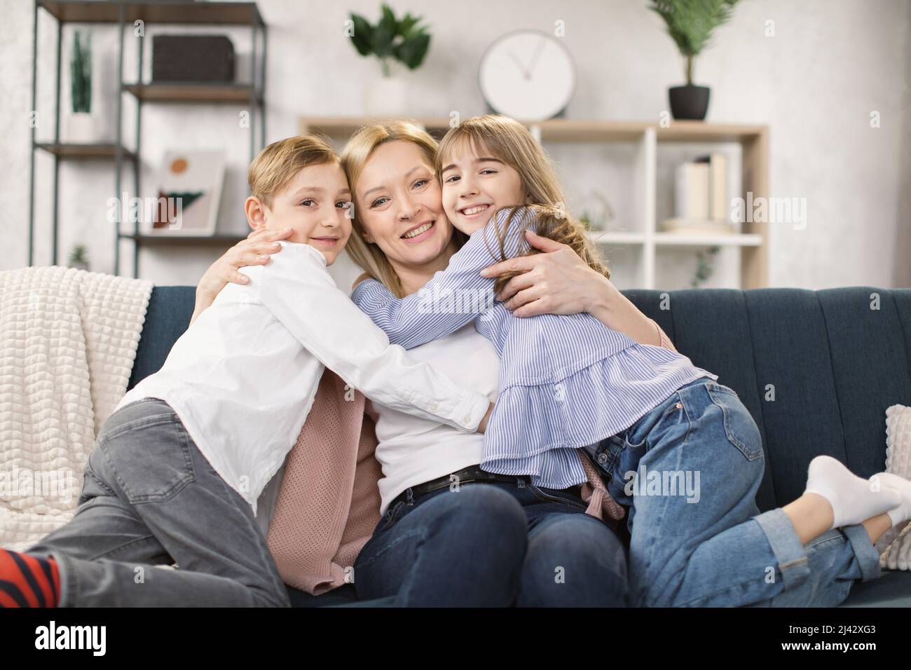 Happy little kids sit on couch hug cuddle excited young mom or nanny ...