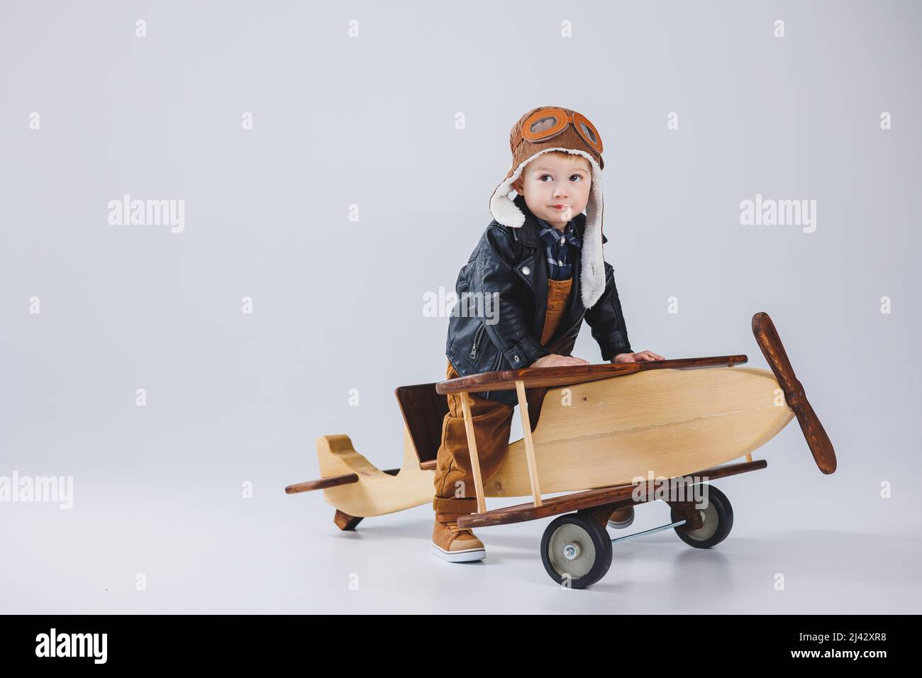 A happy boy in a helmet and a pilot's jacket stands near a wooden plane ...