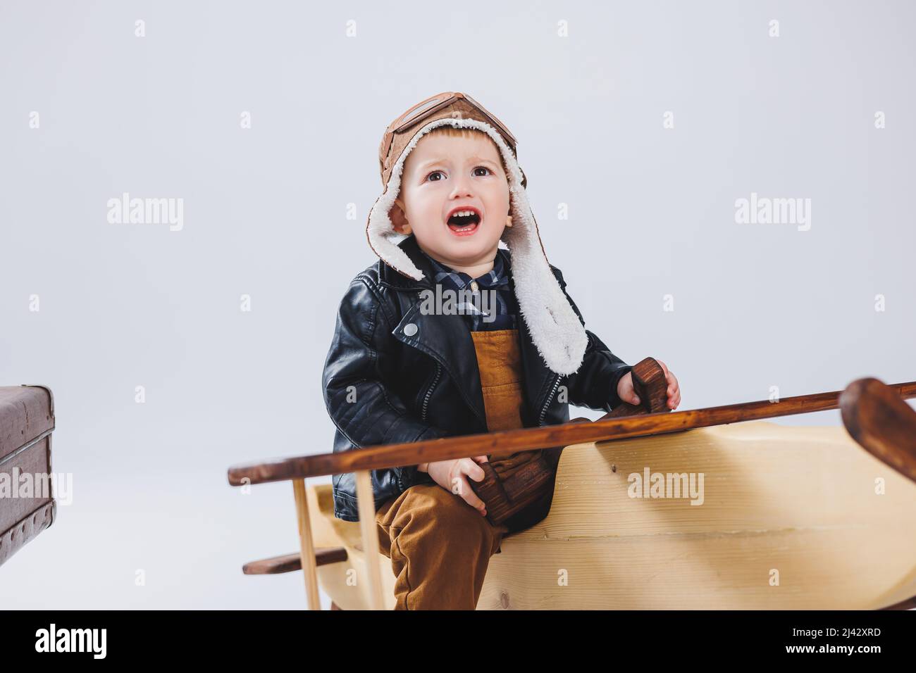 A happy boy in a helmet and a pilot's jacket stands near a wooden plane ...