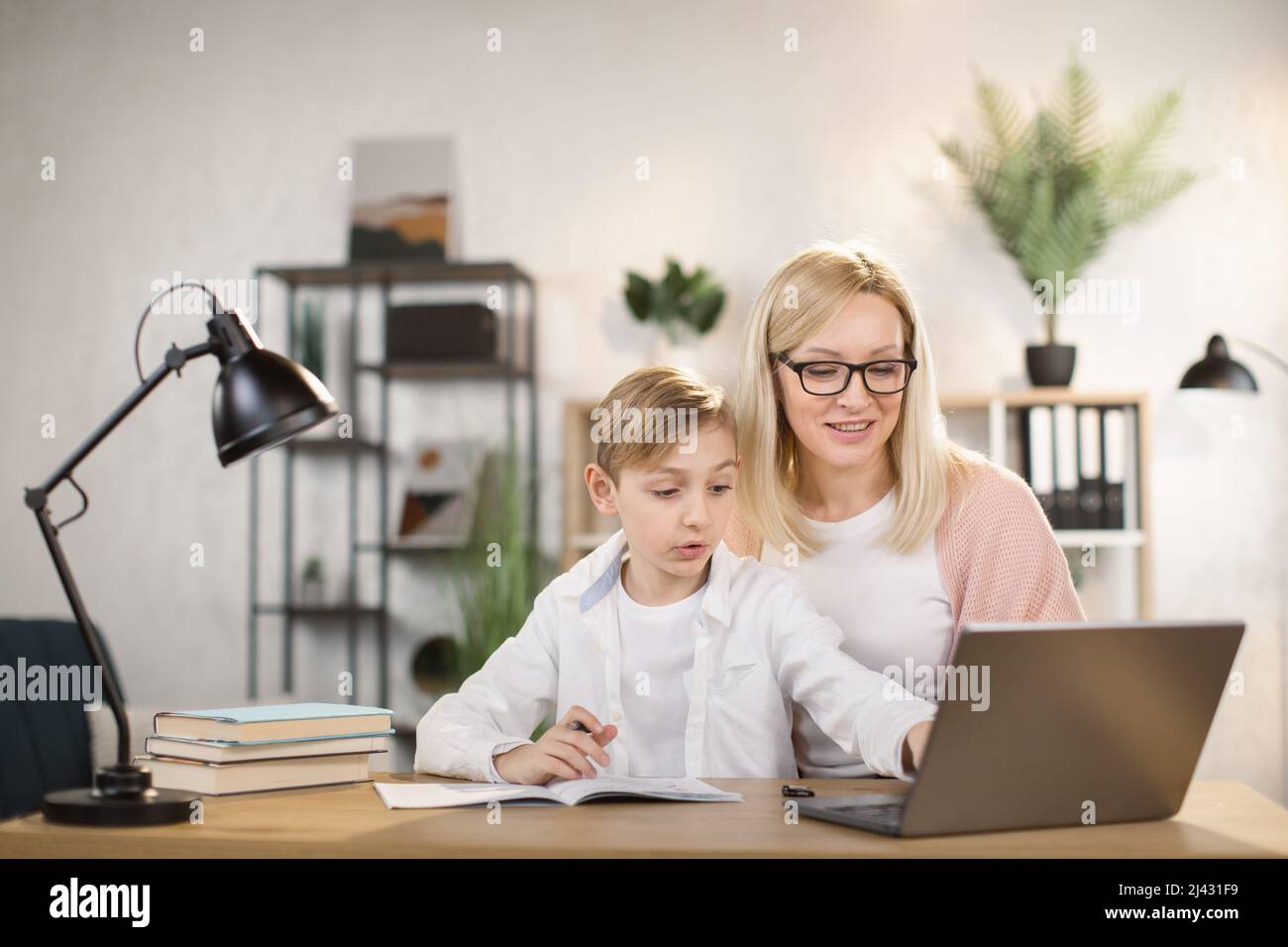 Young mother in eyeglasses pointing at laptop while explaining lesson to little son during ...