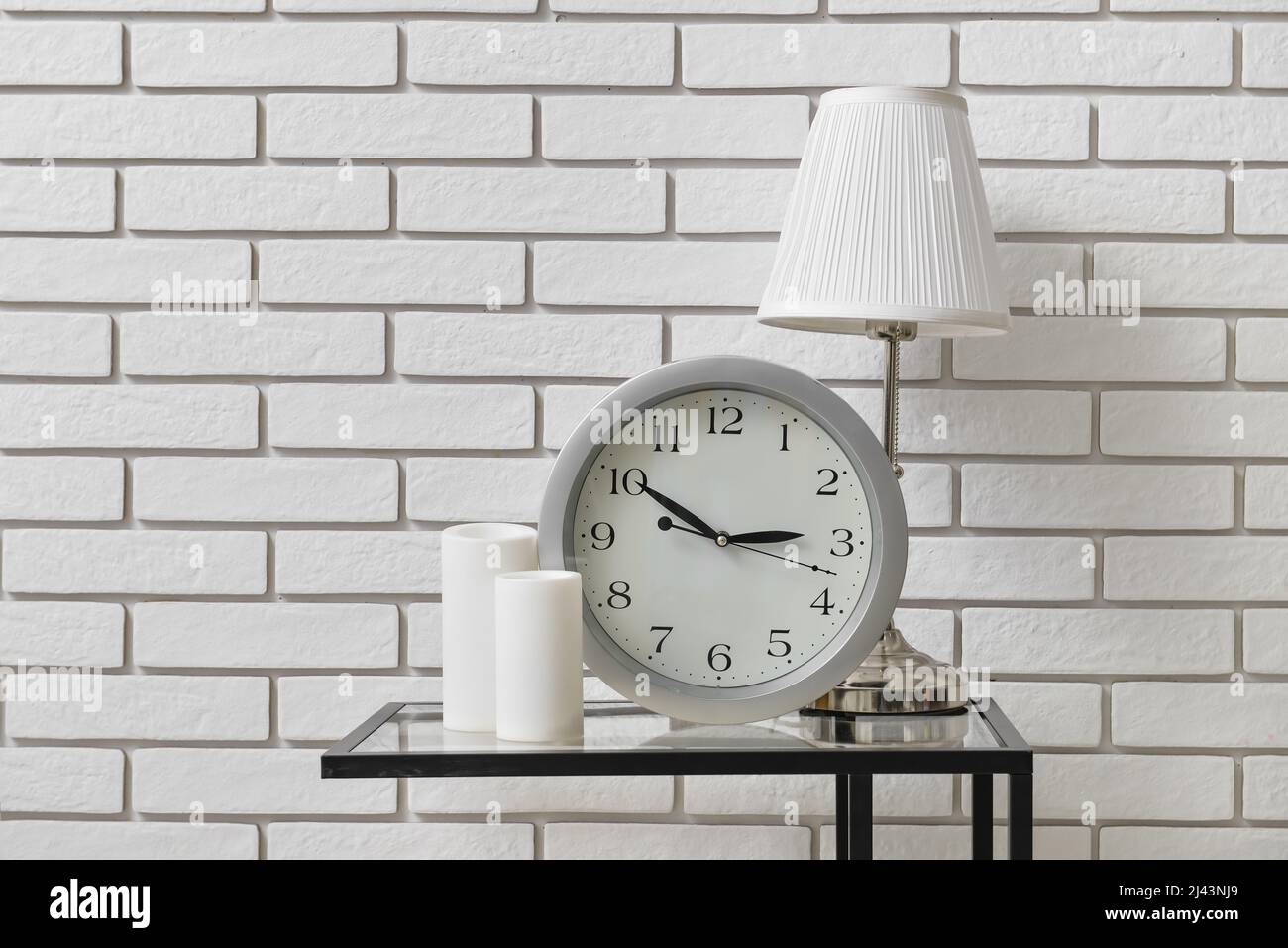 Table with modern clock, lamp and candles near light brick wall in room ...