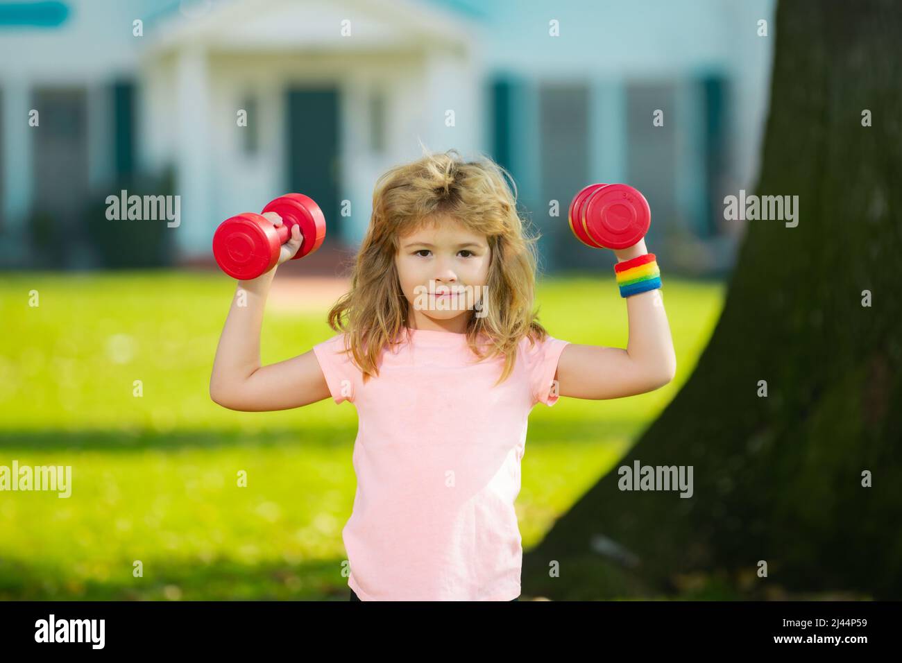 Cute child boy pumping up arm muscles with dumbbell outdoor in summer park. Fitness kids with ...