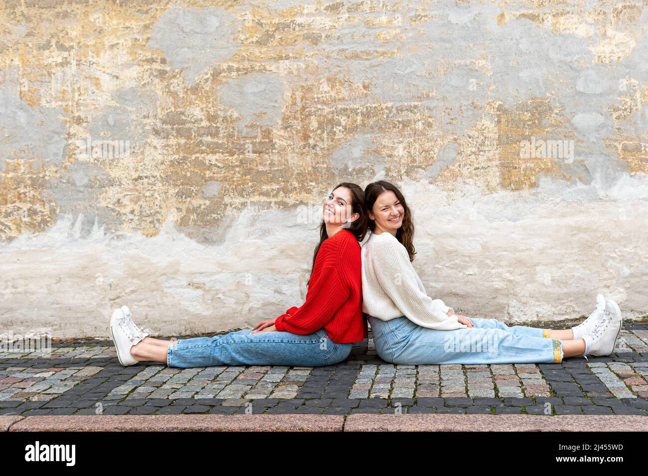 two young women sitting back to back on the pavement by an old wall ...