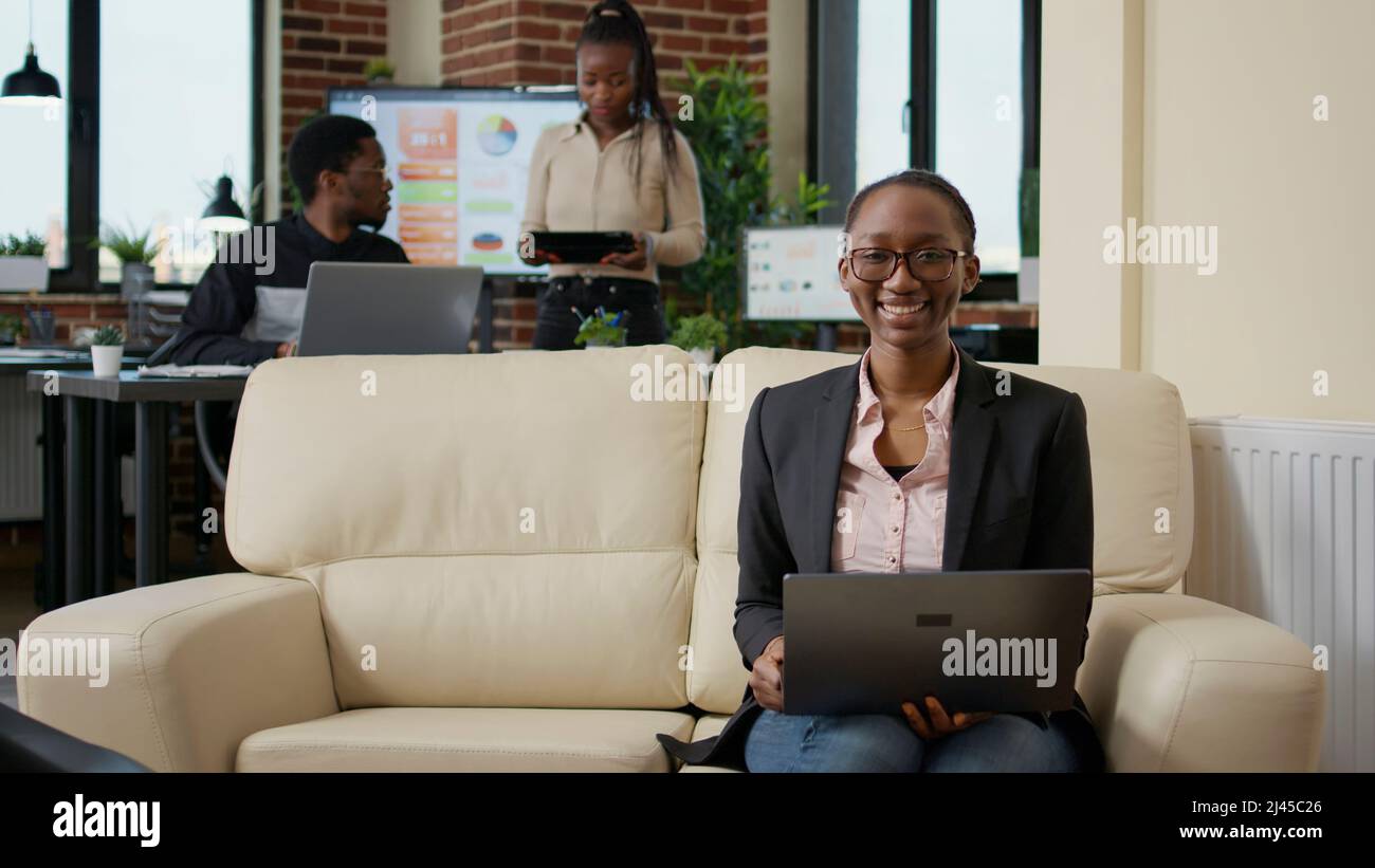 Portrait of african american woman holding laptop on sofa in company ...