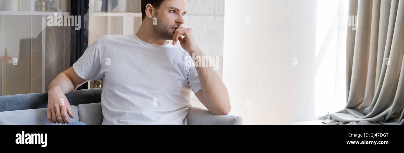 young man with hand near face looking away while thinking at home ...