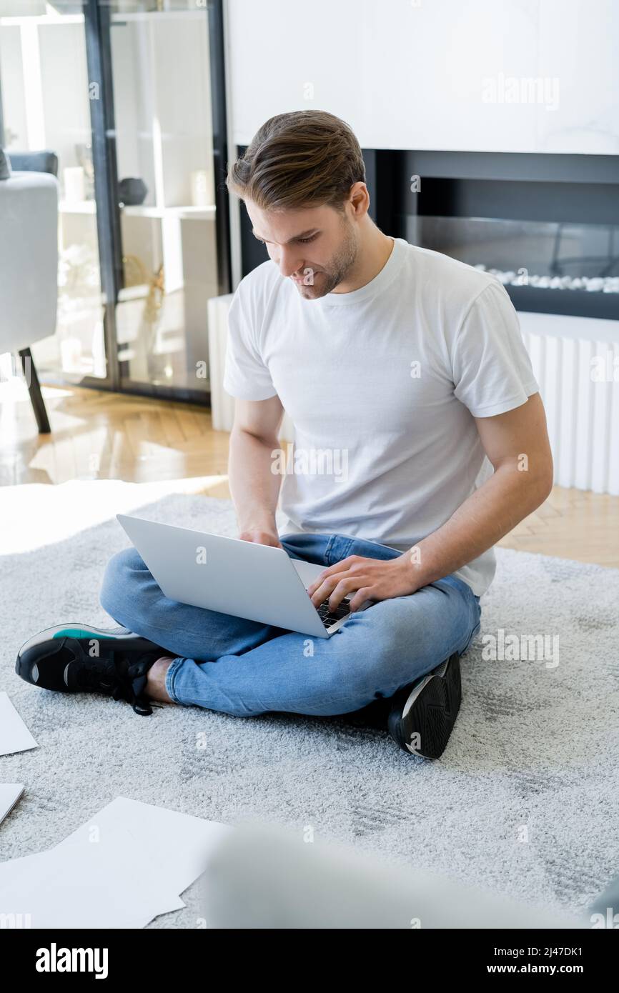 full length of man sitting on floor with crossed legs and typing on ...