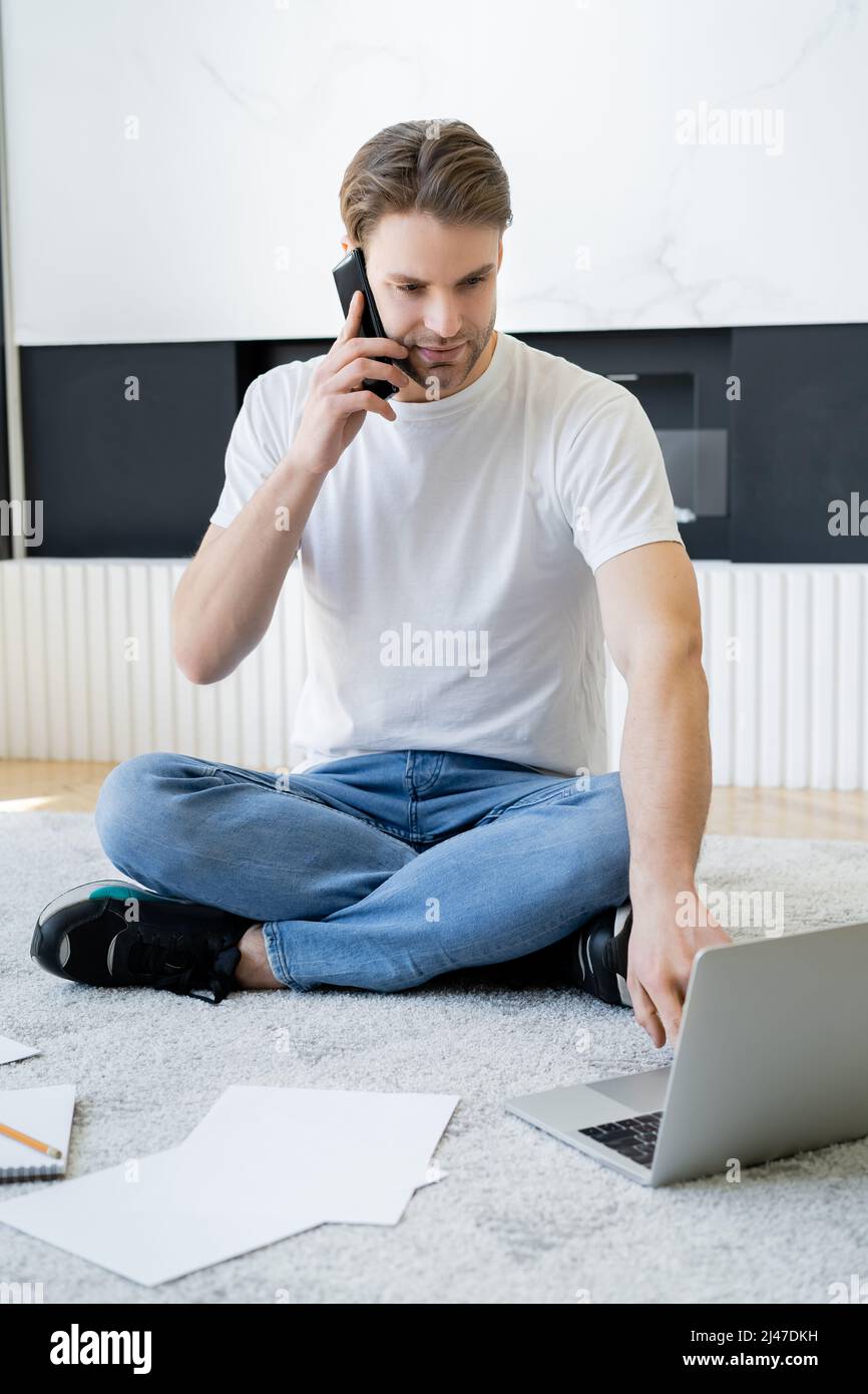 young man sitting on floor with crossed legs and talking on smartphone ...