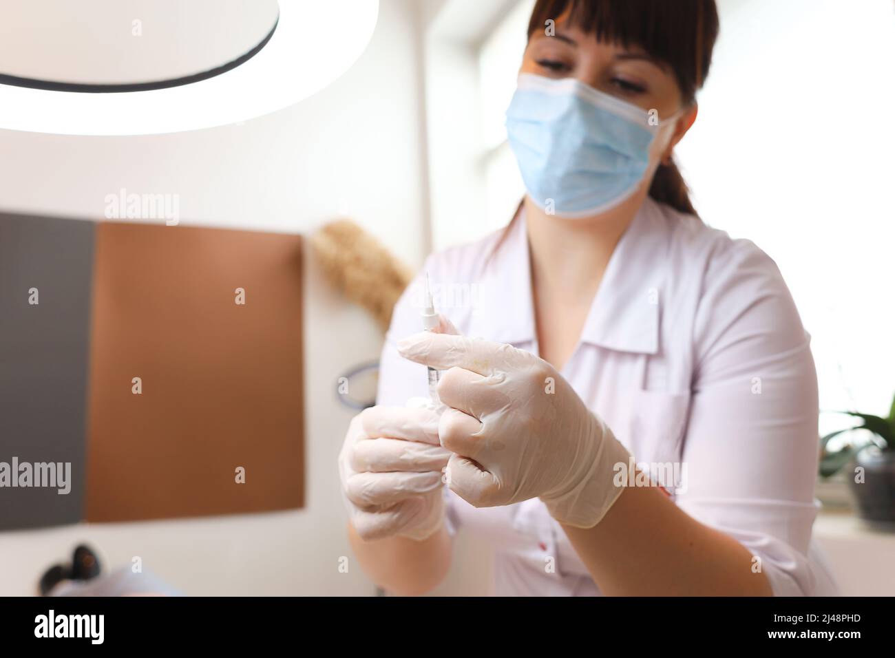 A beautician wearing sterile white gloves holds a hyaluronic acid injection filler and looks at ...