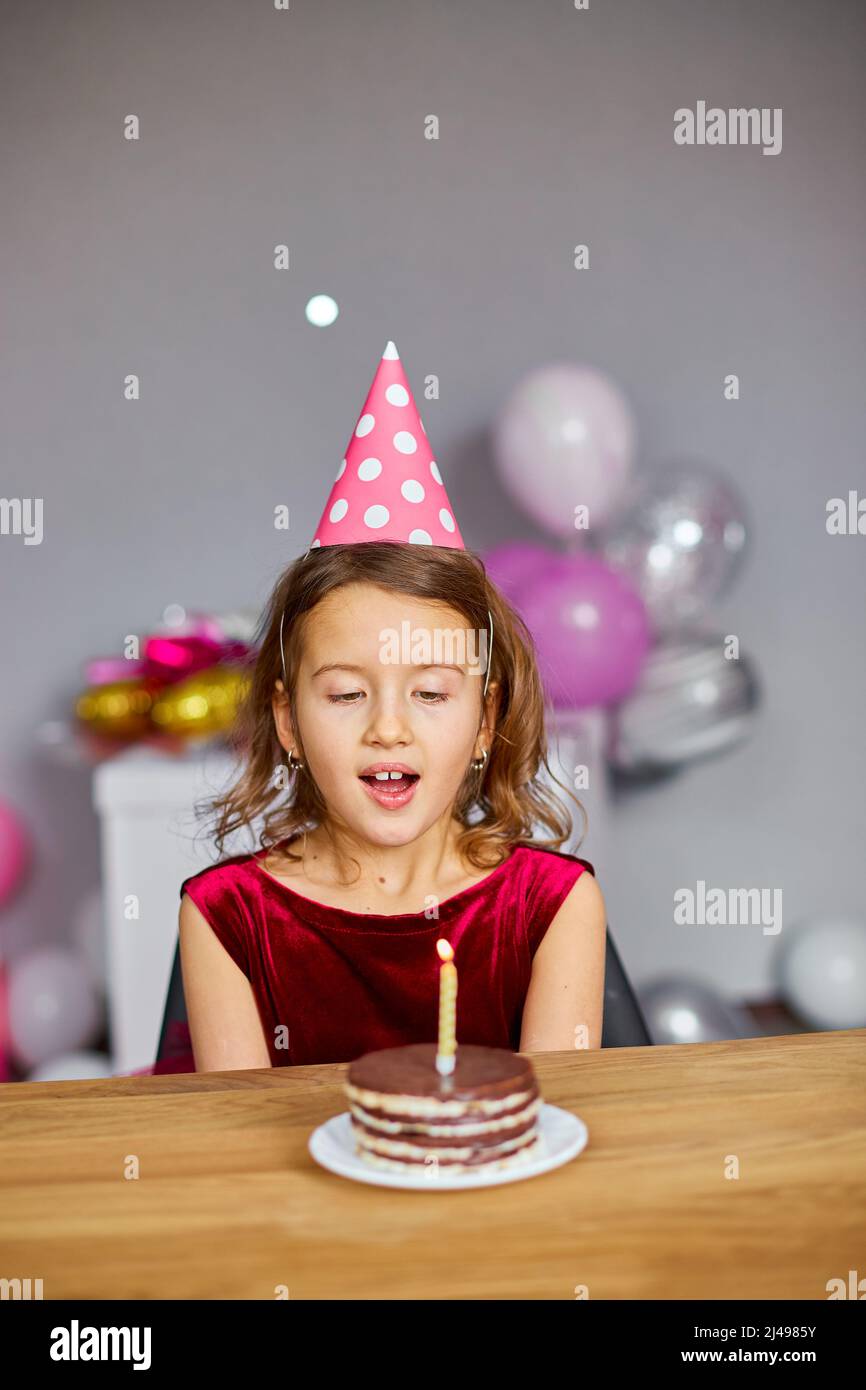 A little girl is wearing a birthday hat looking at a Birthday Cake and ...