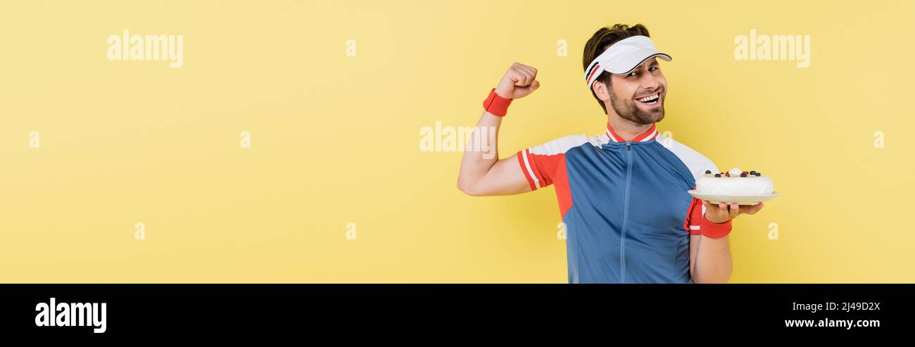 Cheerful sportsman holding cake and showing muscles isolated on yellow ...