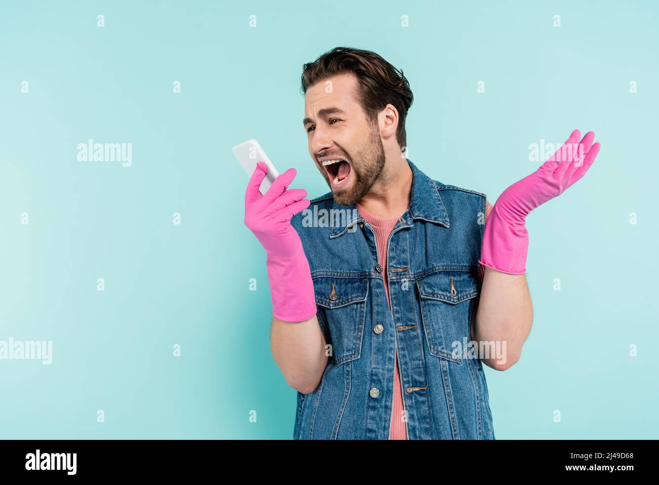 Angry man in rubber gloves looking at smartphone isolated on blue Stock ...