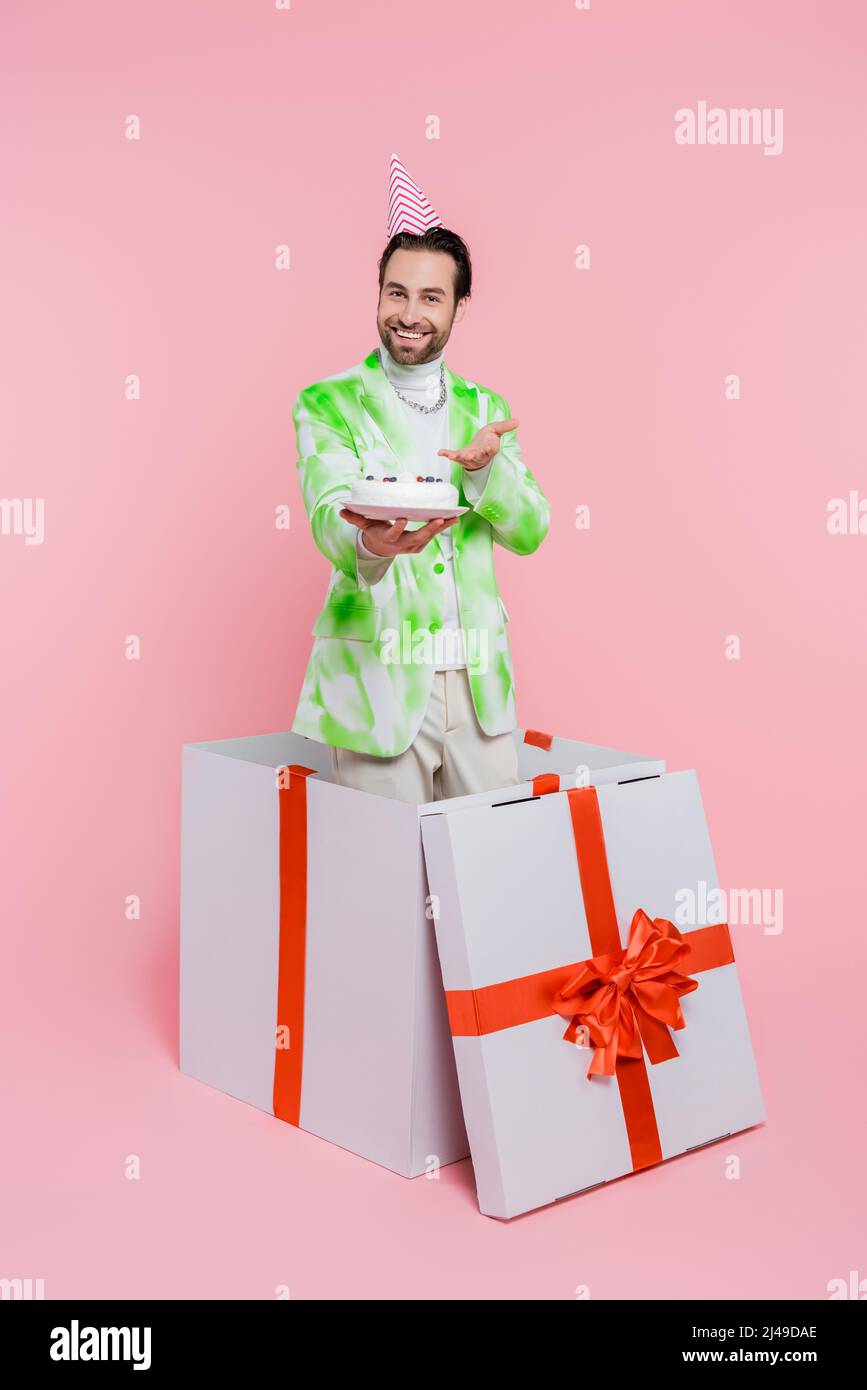 Positive man in party cap pointing at birthday cake in gift box on pink ...
