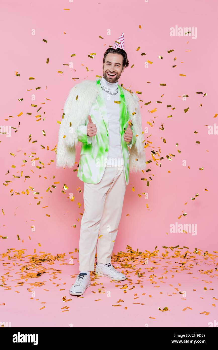 Cheerful man in furry jacket and party cap showing like under confetti ...
