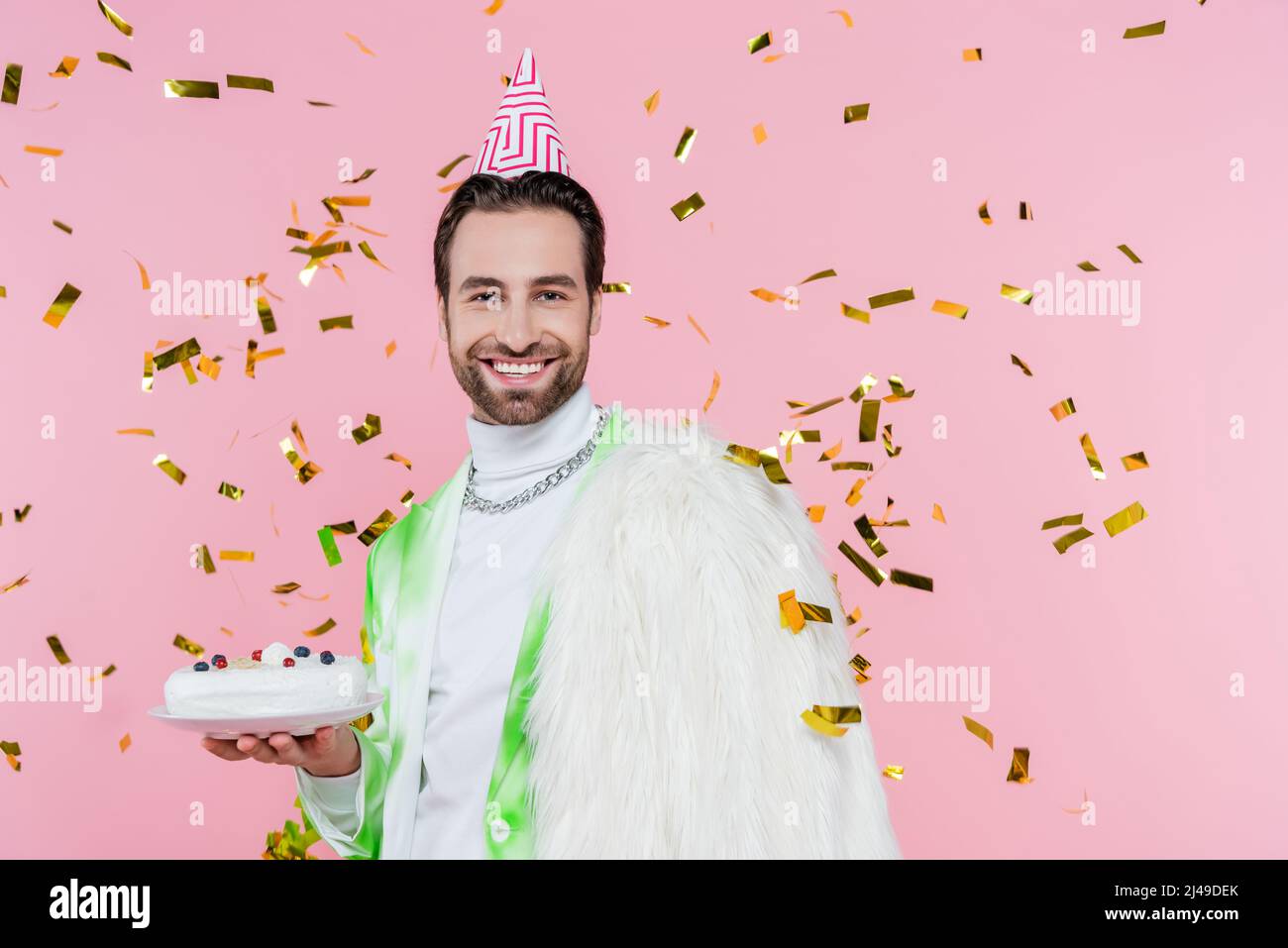Positive man in furry jacket and party cap holding birthday cake under ...