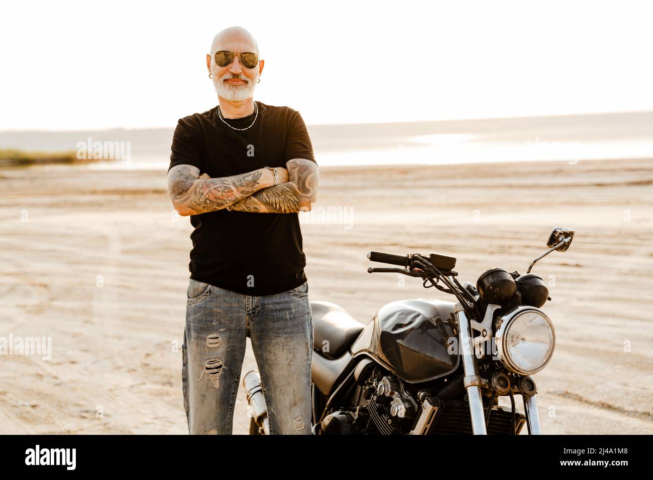 Mature bold man with tattoo looking at camera while posing by motorbike ...