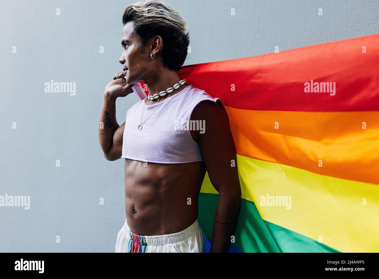 Young gay standing with a pride flag at grey wall and looking away ...