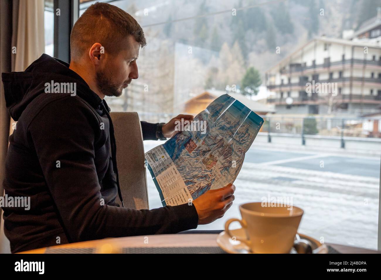 Young man reading paper map while sitting beside table with cup at ...