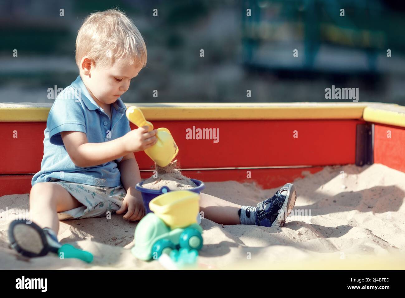 Toddler boy is playing in a red and yellow edge sandbox with sand toys ...