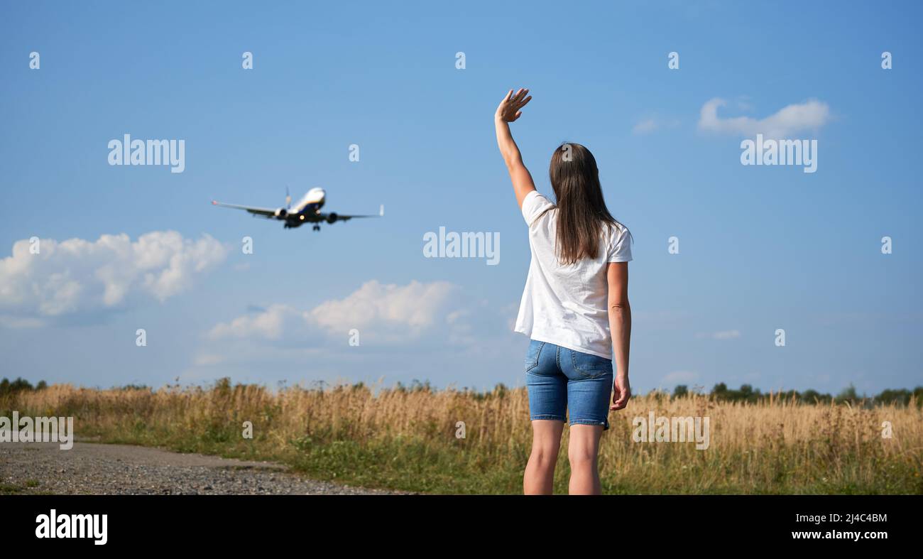Back view of woman waving hand to flying commercial airplane in the sky ...