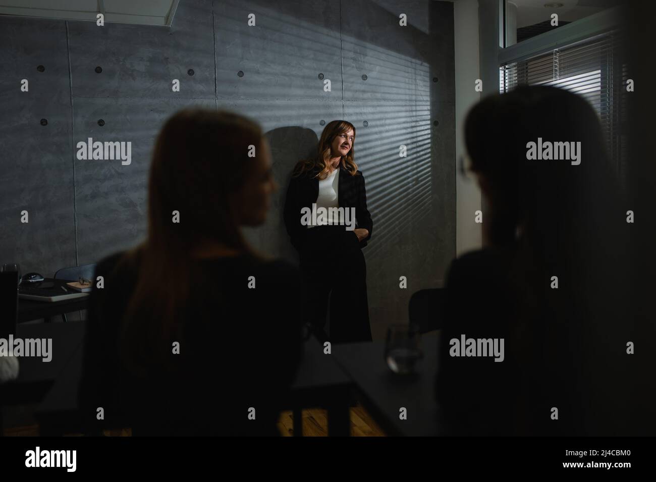 A senior, smiling female teacher leans against a gray wall in a ...