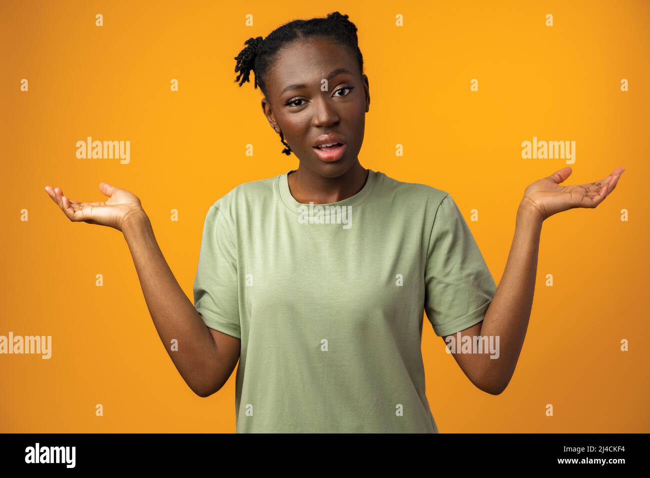 Puzzled and clueless young African woman with arms out in yellow studio ...